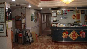 Uma área de recepção de hotel com piso de mármore, balcão de atendimento decorado, um lustre e diversas decorações nas paredes, incluindo calendários e quadros.