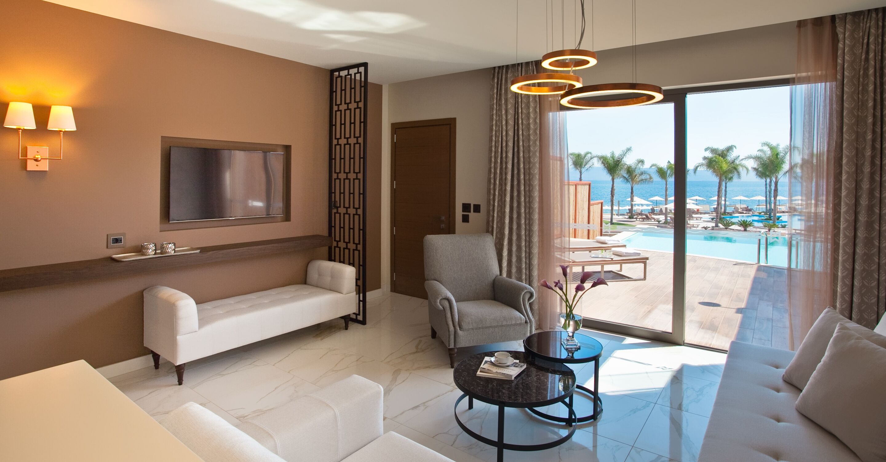 Quarto de hotel moderno com sofá, poltrona e mesa de centro. Uma televisão está fixada na parede, e há uma grande janela com vista para uma piscina e palmeiras.