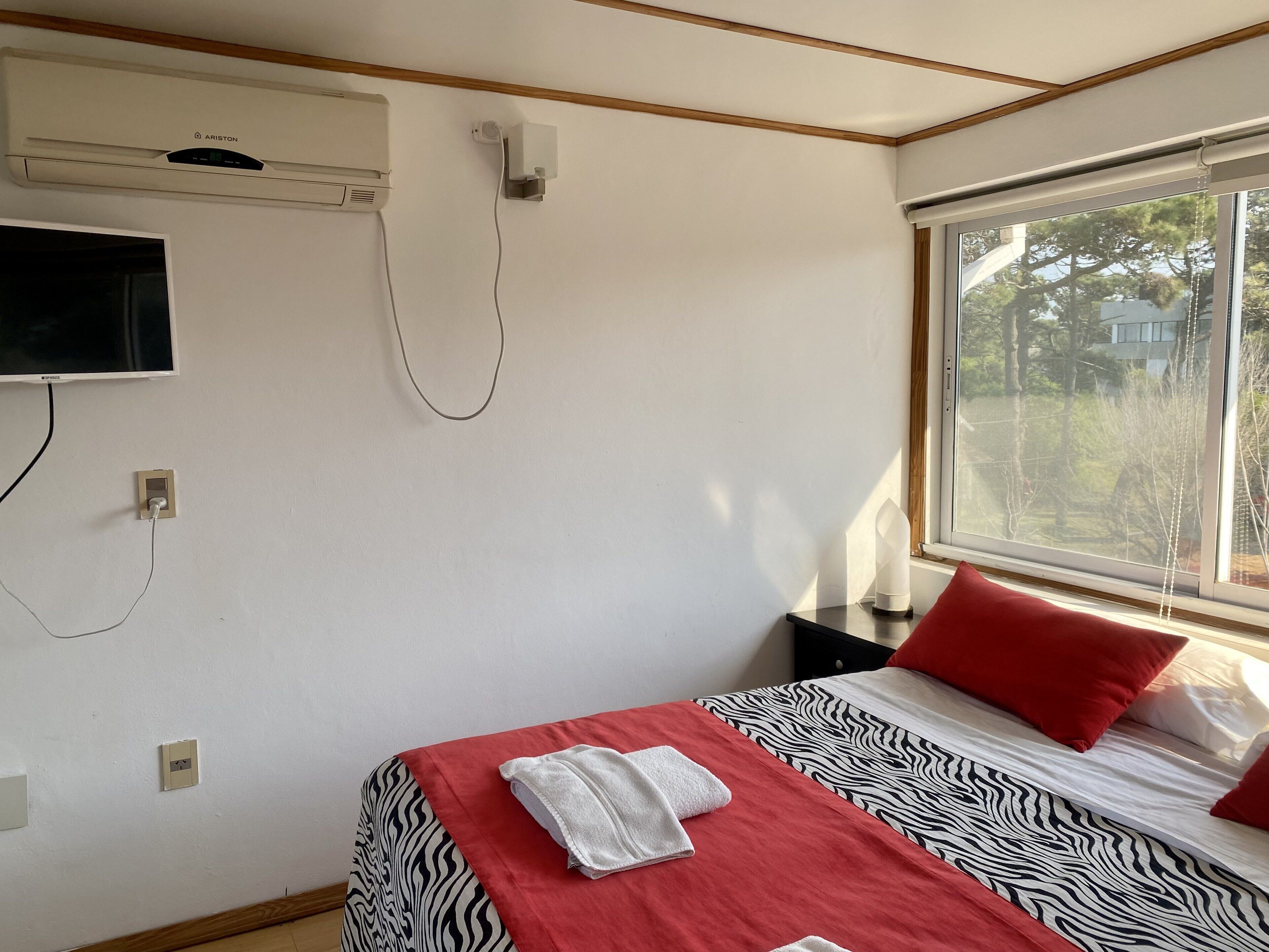 Quarto de hotel com uma cama, TV fixada na parede e janela com vista para árvores.