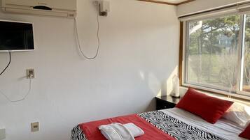 Quarto de hotel com uma cama, TV fixada na parede e janela com vista para árvores.