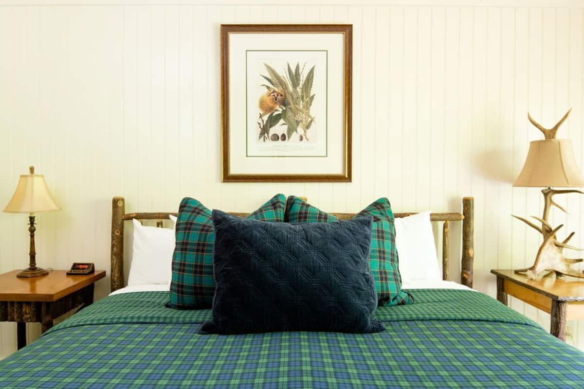 Uma cama bem arrumada com colcha verde xadrez, dois travesseiros, uma ilustração botânica emoldurada e uma mesa de cabeceira de madeira com uma luminária.