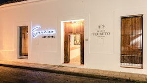 Um edifício branco com um letreiro de neon para 'Sereno Art Hotel' e uma porta de madeira com a inscrição 'Jardín Secretó Cocina Artesanal'.