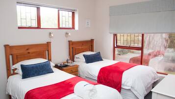 Quarto de hotel com duas camas, cada uma com cabeceira de madeira, roupa de cama branca e almofadas azuis e vermelhas. Tem uma pequena mesa de cabeceira com uma luminária e uma janela com vista para o exterior.
