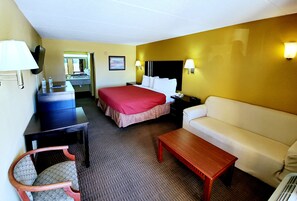 Quarto de hotel com cama, sofá, mesa de madeira, cadeira, escrivaninha com luminária, TV e banheiro visível ao fundo.