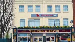 Um edifício com a placa do pub 'The Pick & Shovel', a placa 'ELSTREE INN' e uma placa com a inscrição 'COZINHA NOVA ABERTA'.