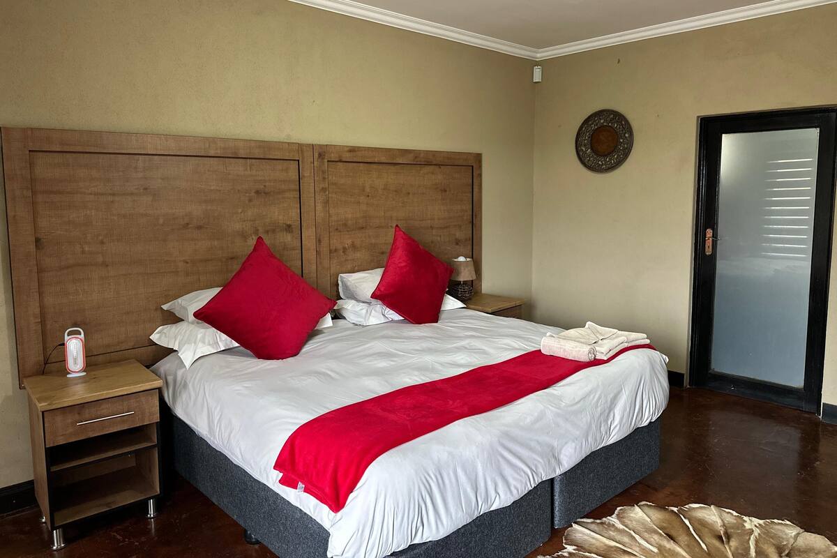 Quarto de hotel com uma cama grande, duas mesas de cabeceira, cabeceira de madeira, manta vermelha e uma porta com vista.