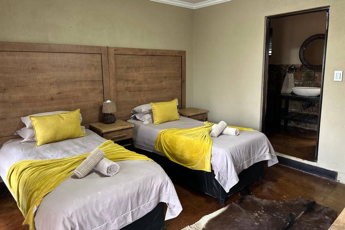 Quarto de hotel com duas camas, cada uma com um cobertor amarelo e uma toalha enrolada. Há uma cabeceira de madeira, uma mesa de cabeceira com uma luminária, e um banheiro visível através de uma porta aberta.