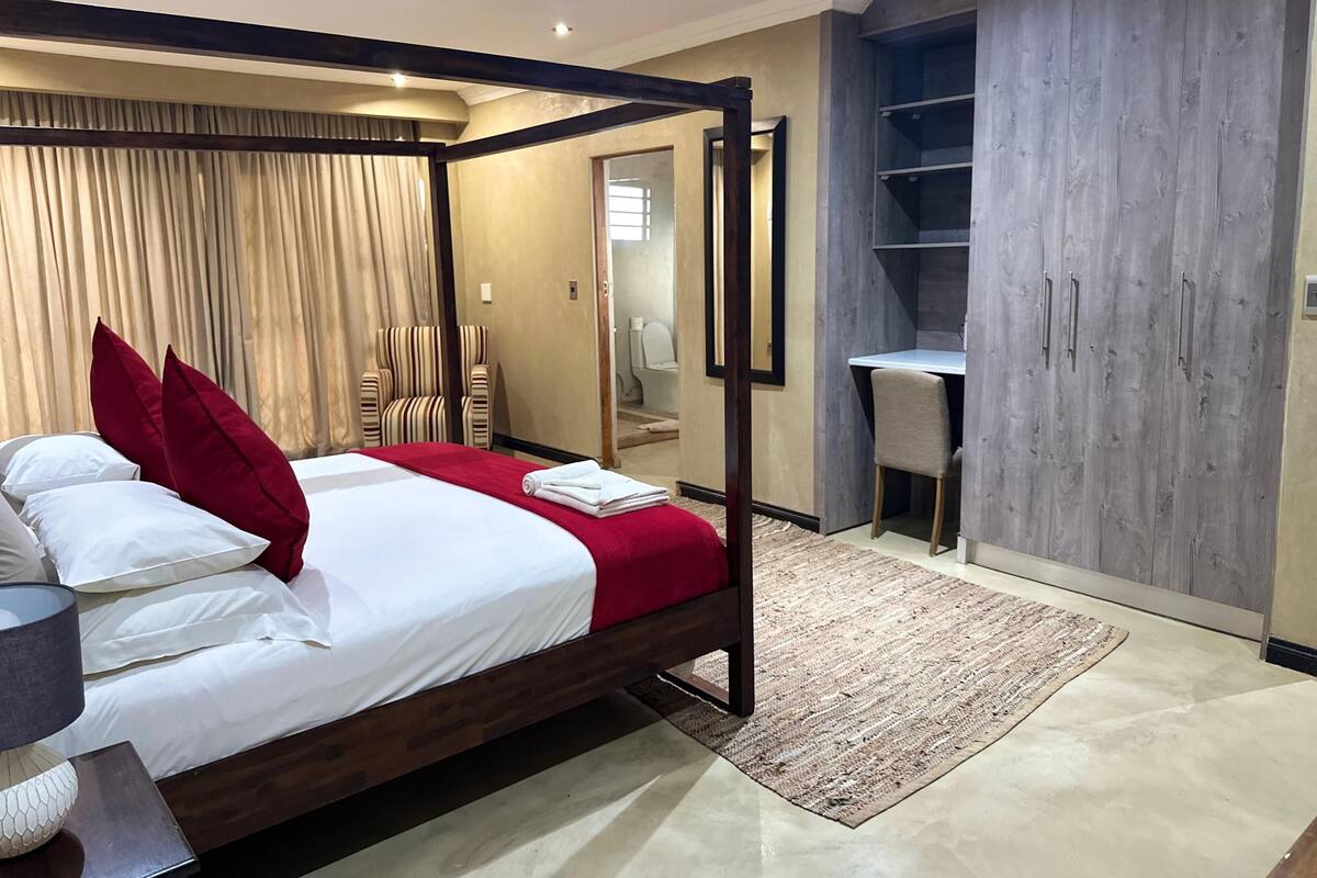 Uma cama com dossel, roupa de cama branca e vermelha, uma mesinha de cabeceira de madeira, uma poltrona listrada, um guarda-roupa grande e um espelho.