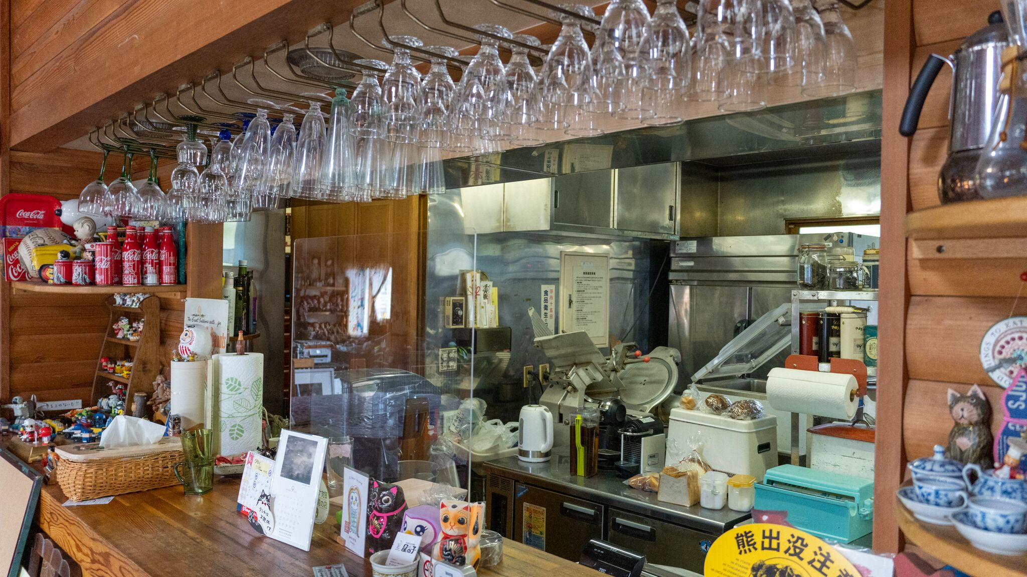 Uma cozinha com diversos eletrodomésticos e utensílios, uma exposição de copos e pratos, e prateleiras com itens como latas de Coca-Cola e figuras decorativas.
