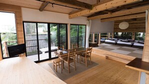 Um quarto espaçoso com piso de madeira, área de jantar com mesa e cadeiras, sofá com almofadas e grandes portas de vidro deslizantes que dão para uma área externa.