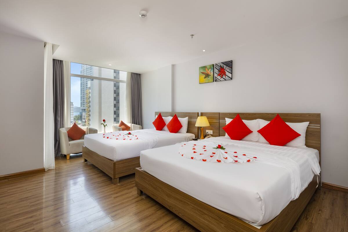 Quarto de hotel com duas camas, cada uma com roupa de cama branca e almofadas vermelhas, cabeceira de madeira, mesa de cabeceira com abajur e uma janela grande com cortinas.