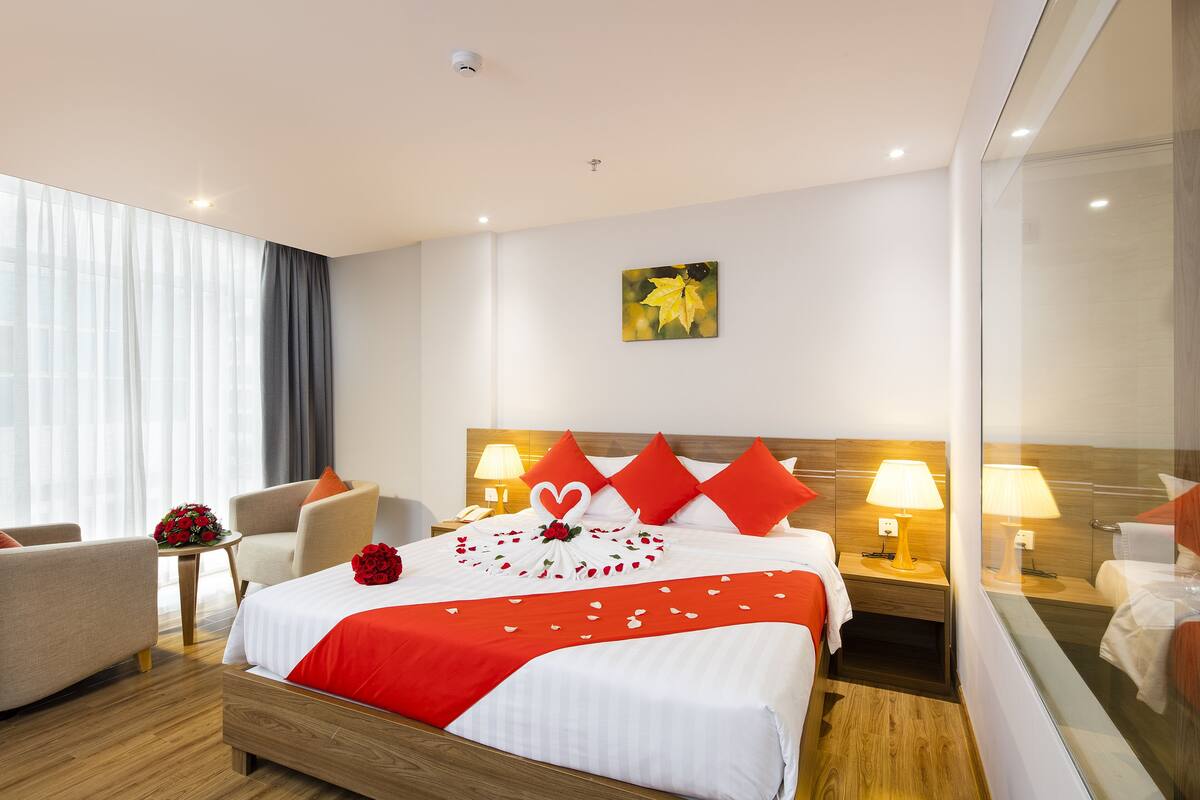 Quarto de hotel com uma cama grande, duas mesas de cabeceira com luminárias, um sofá e uma pequena mesa com um arranjo de flores.