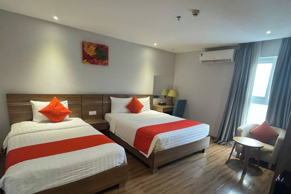 Quarto de hotel com duas camas, uma mesinha, uma cadeira, aparelho de ar condicionado e uma janela com cortinas.