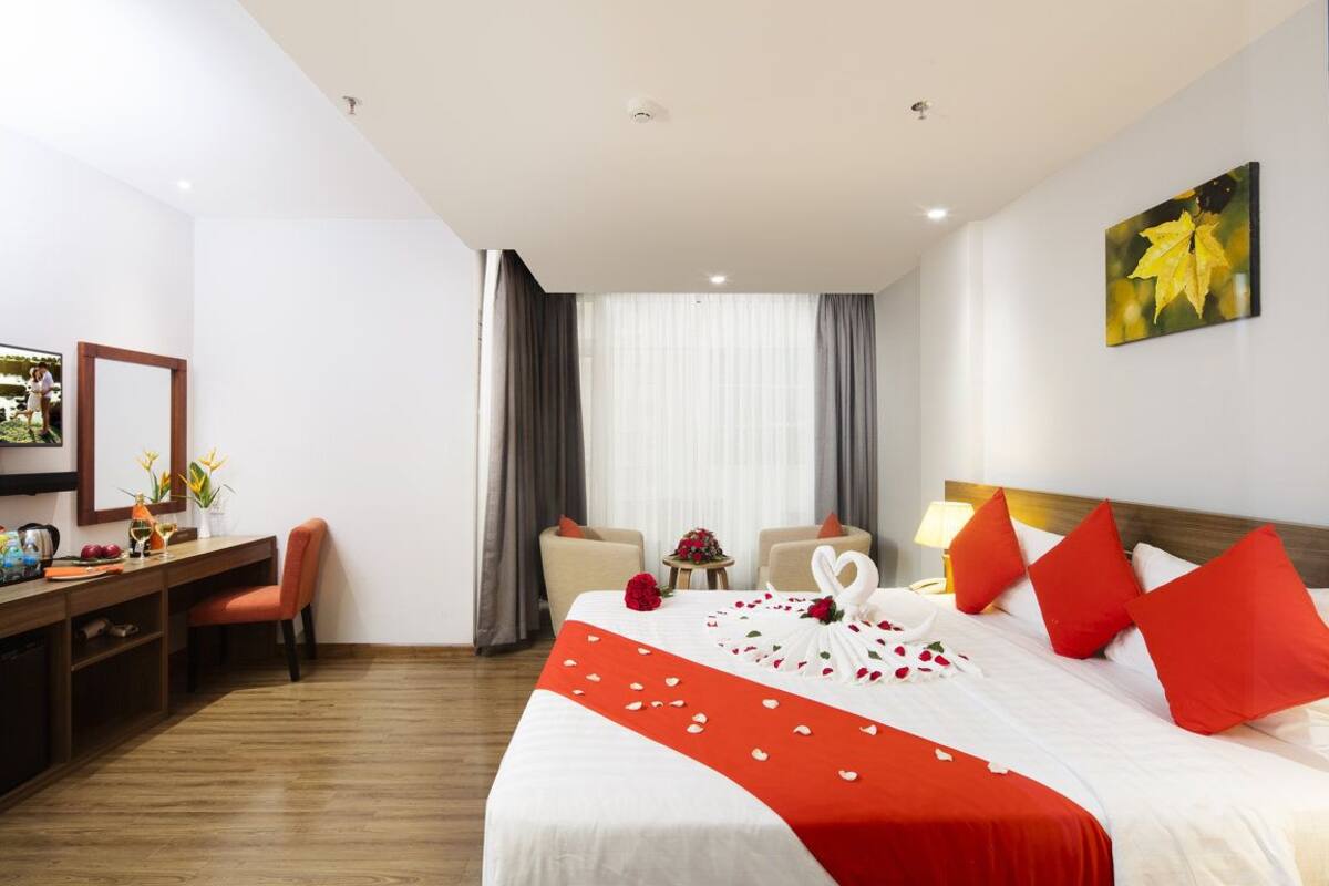 Quarto de hotel com uma cama, uma escrivaninha, uma cadeira, uma mesinha de centro com um vaso e um quadro na parede.