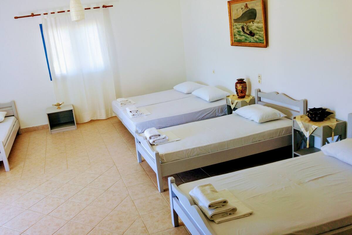 Quarto de hotel com três camas de solteiro, uma mesinha com um vaso e um quadro na parede.
