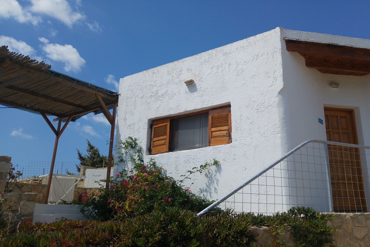 Um edifício branco com porta e venezianas de madeira, um alpendre coberto e uma parede de pedra.