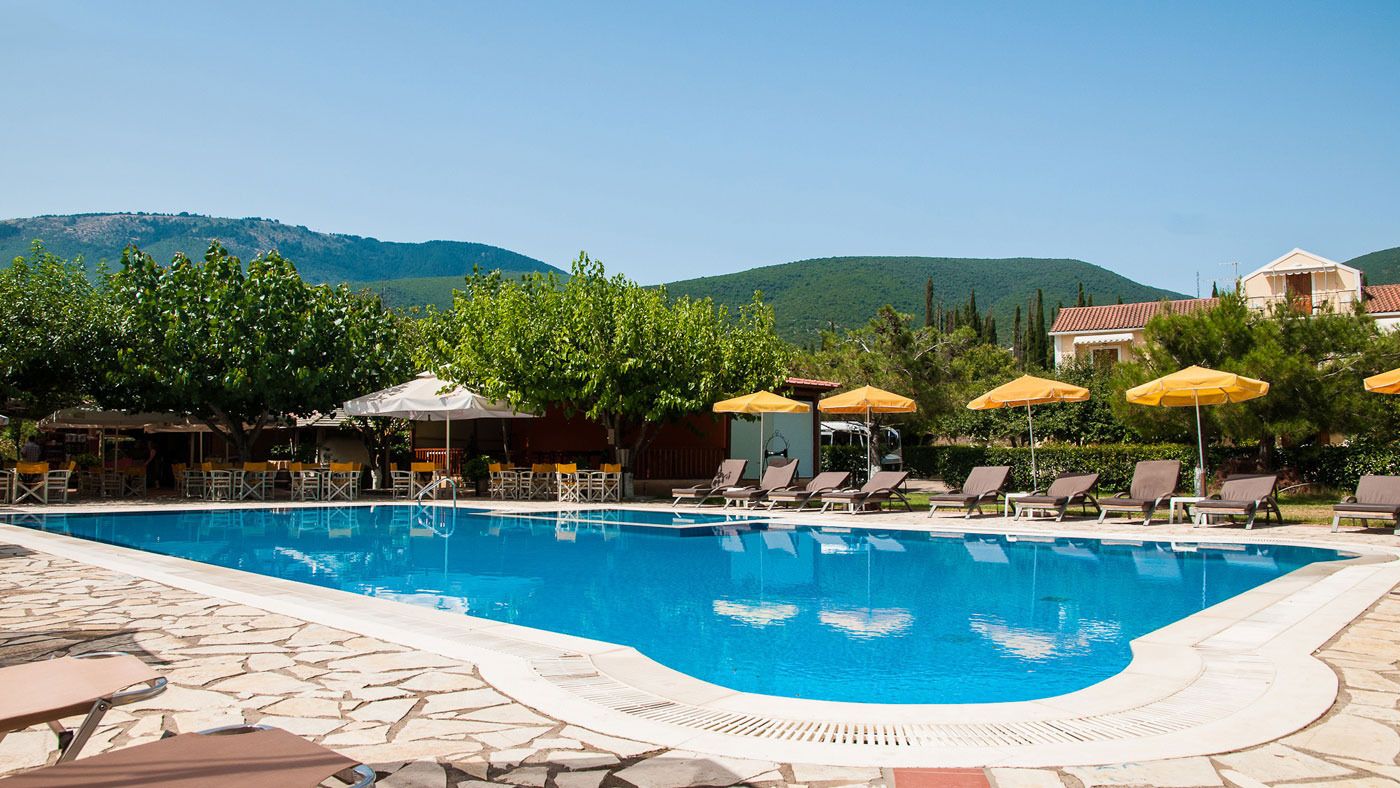 Piscina com espreguiçadeiras e guarda-sóis, rodeada por árvores e edifícios, com vista para colinas ao fundo.