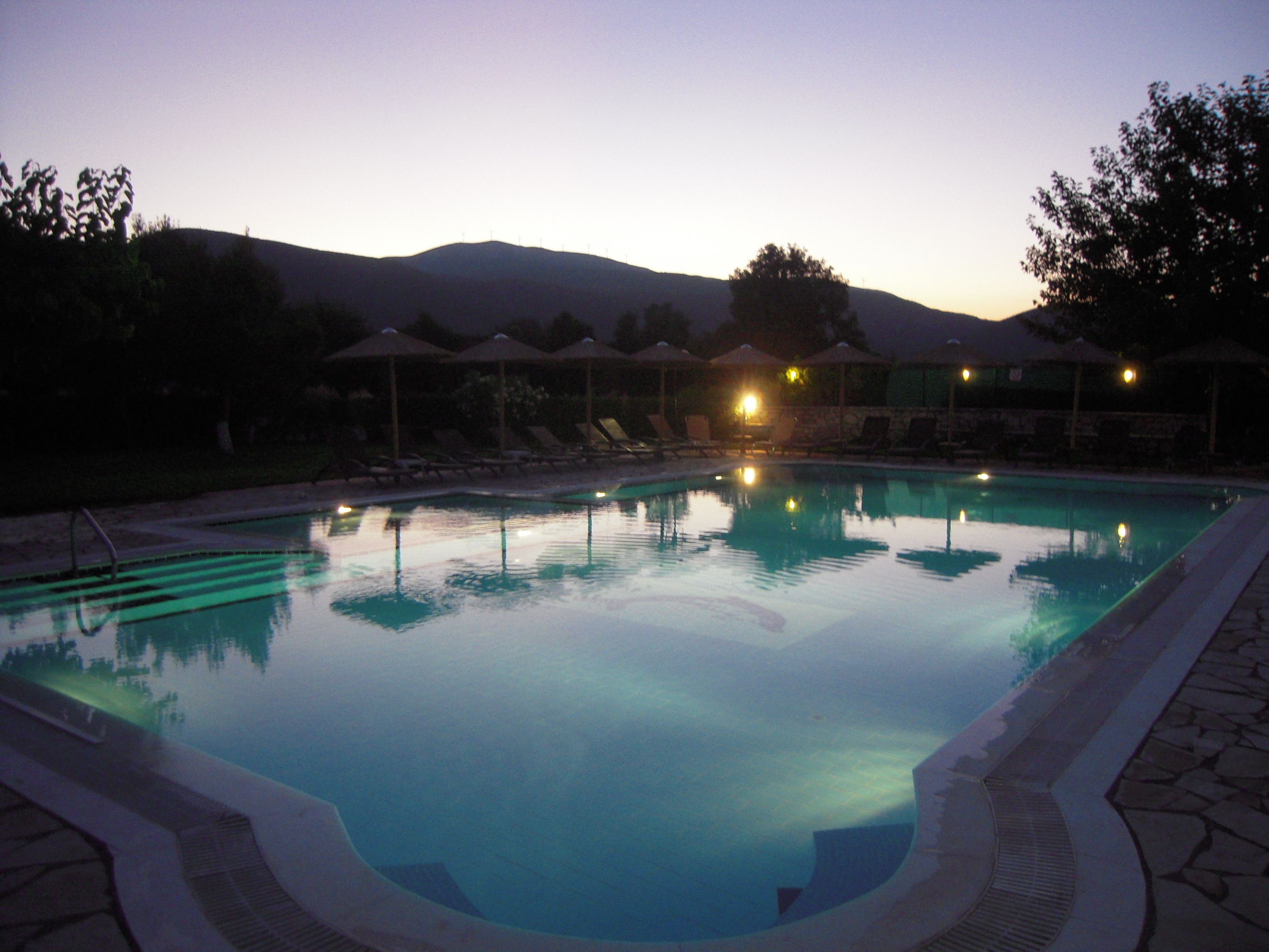 Uma piscina com espreguiçadeiras e guarda-sóis, rodeada por árvores e colinas.
