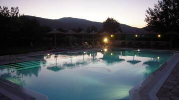 Uma piscina com espreguiçadeiras e guarda-sóis, rodeada por árvores e colinas.