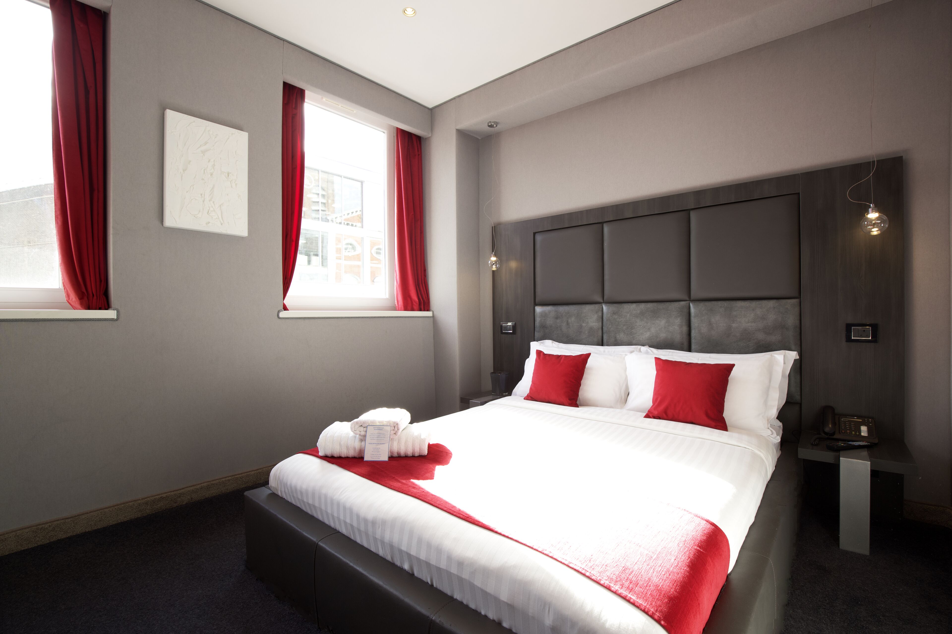 Quarto de hotel moderno com uma cama grande, cortinas vermelhas, cabeceira com luminária e mesa de cabeceira com telefone.