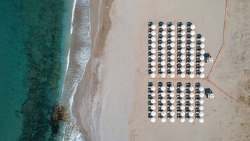 Vista aérea de uma praia com fileiras de guarda-sóis e cadeiras, indicando um resort à beira-mar.