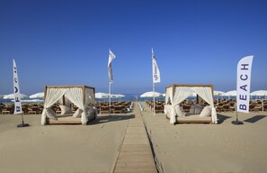Um resort à beira-mar com duas camas de praia cobertas por toldos, um deck de madeira e bandeiras com a palavra 'BEACH'.