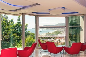 Quarto moderno com grandes janelas que oferecem vista da cidade, cadeiras vermelhas e divisória de vidro.