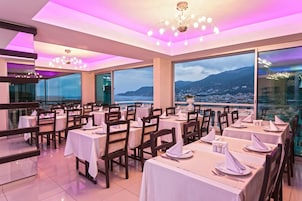 Restaurante com várias mesas preparadas para refeições, grandes janelas com vista para uma cidade e montanhas, e um lustre moderno.