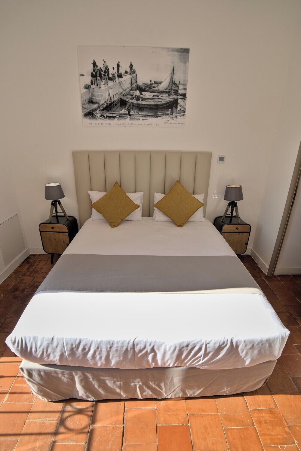 Uma cama com cabeceira, duas almofadas, uma mesa de cabeceira com abajur e um quadro na parede.