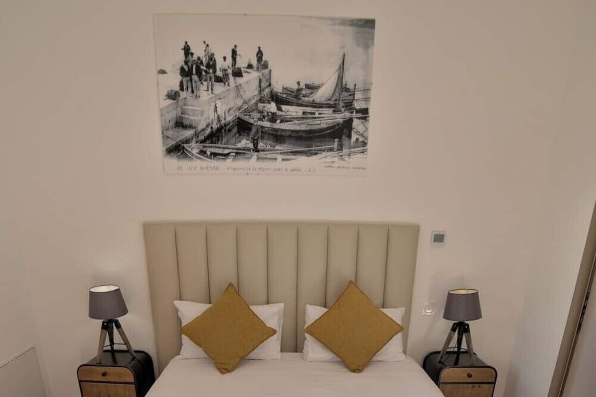 Uma cama com cabeceira, duas almofadas, uma mesa de cabeceira com abajur e um quadro na parede.
