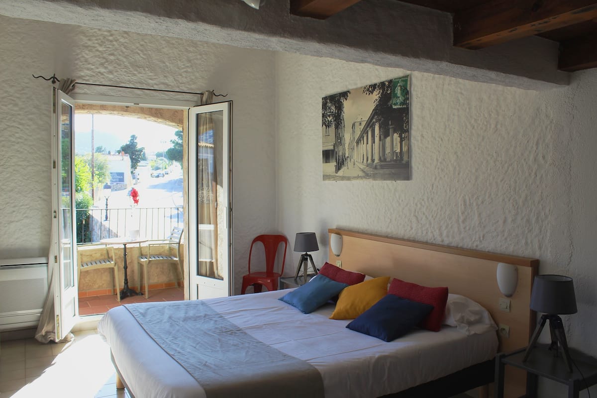 Quarto com cama, mesa de cabeceira, luminária, cadeira e varanda com vista para a rua.