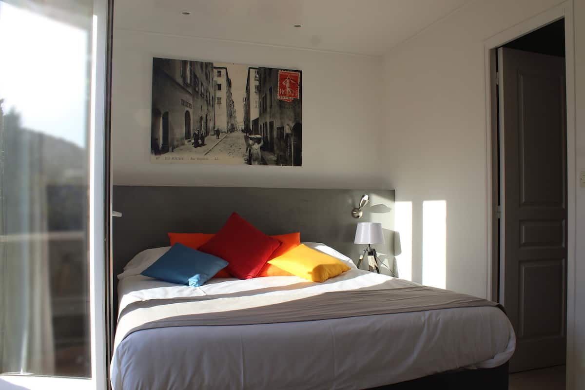 Uma cama bem arrumada com almofadas coloridas, uma luminária na parede e um quadro emoldurado em cima.