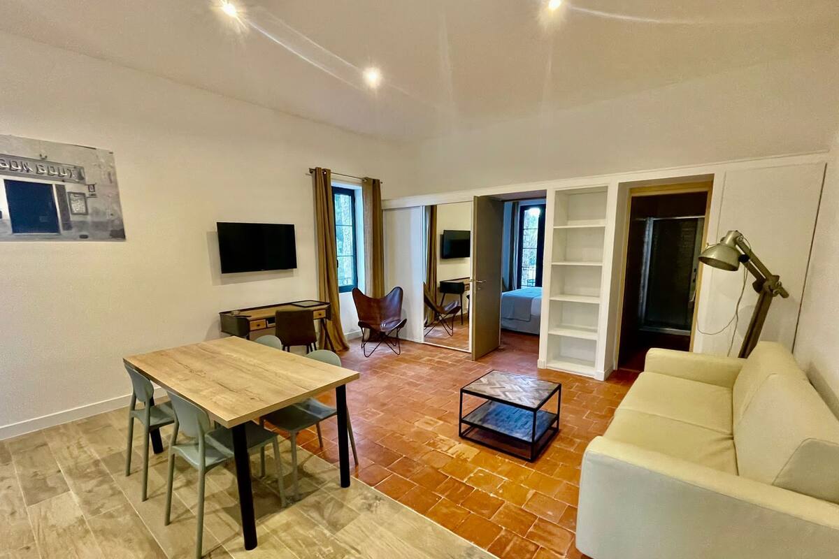 Uma sala de estar ampla com uma mesa de jantar de madeira, um sofá branco e uma TV fixada na parede.