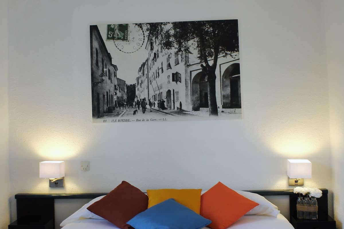 Cama bem arrumada com edredom branco e almofadas coloridas, um quadro com pintura de paisagem urbana em preto e branco acima dela e duas luminárias de cabeceira em mesas de cabeceira pretas.