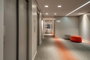 Corredor moderno de hotel, com design alongado, iluminação embutida e piso coberto de carpete.