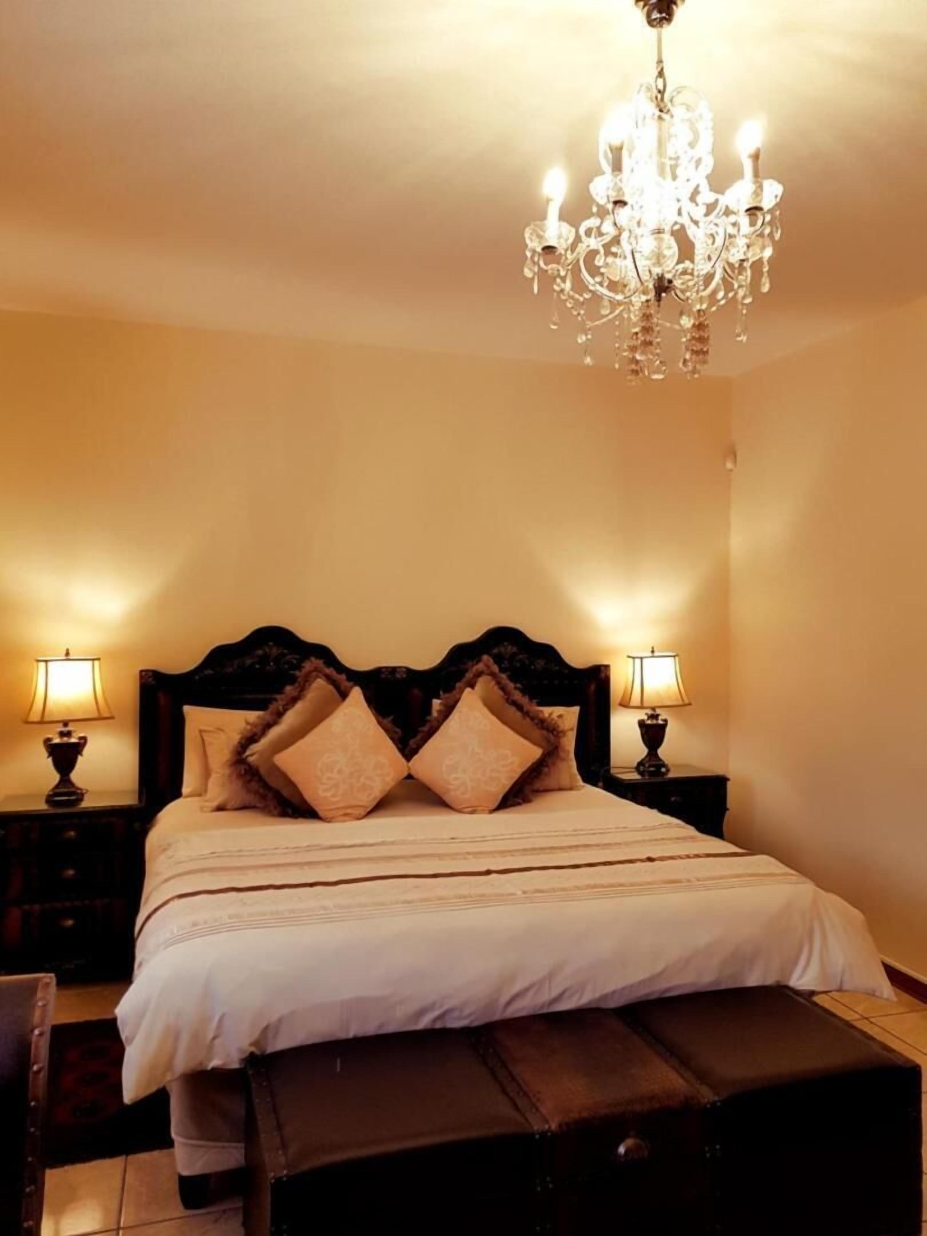 Quarto de hotel com uma cama grande, duas mesas de cabeceira com abajures, um painel de cama de madeira escura e um lustre.