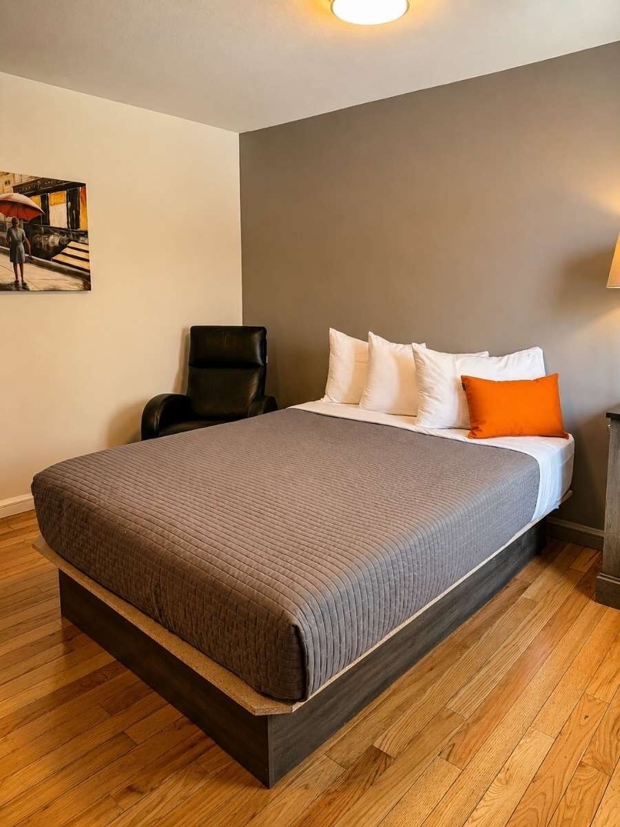 Quarto de hotel com uma cama grande, uma poltrona, uma mesa de cabeceira e um quadro na parede.