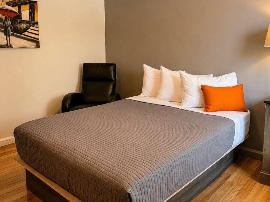 Quarto de hotel com uma cama grande, uma poltrona, uma mesa de cabeceira e um quadro na parede.