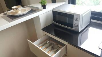 Uma cozinha compacta com micro-ondas, uma gaveta com utensílios, uma chávena e pires numa bancada, e uma pequena planta em vaso.