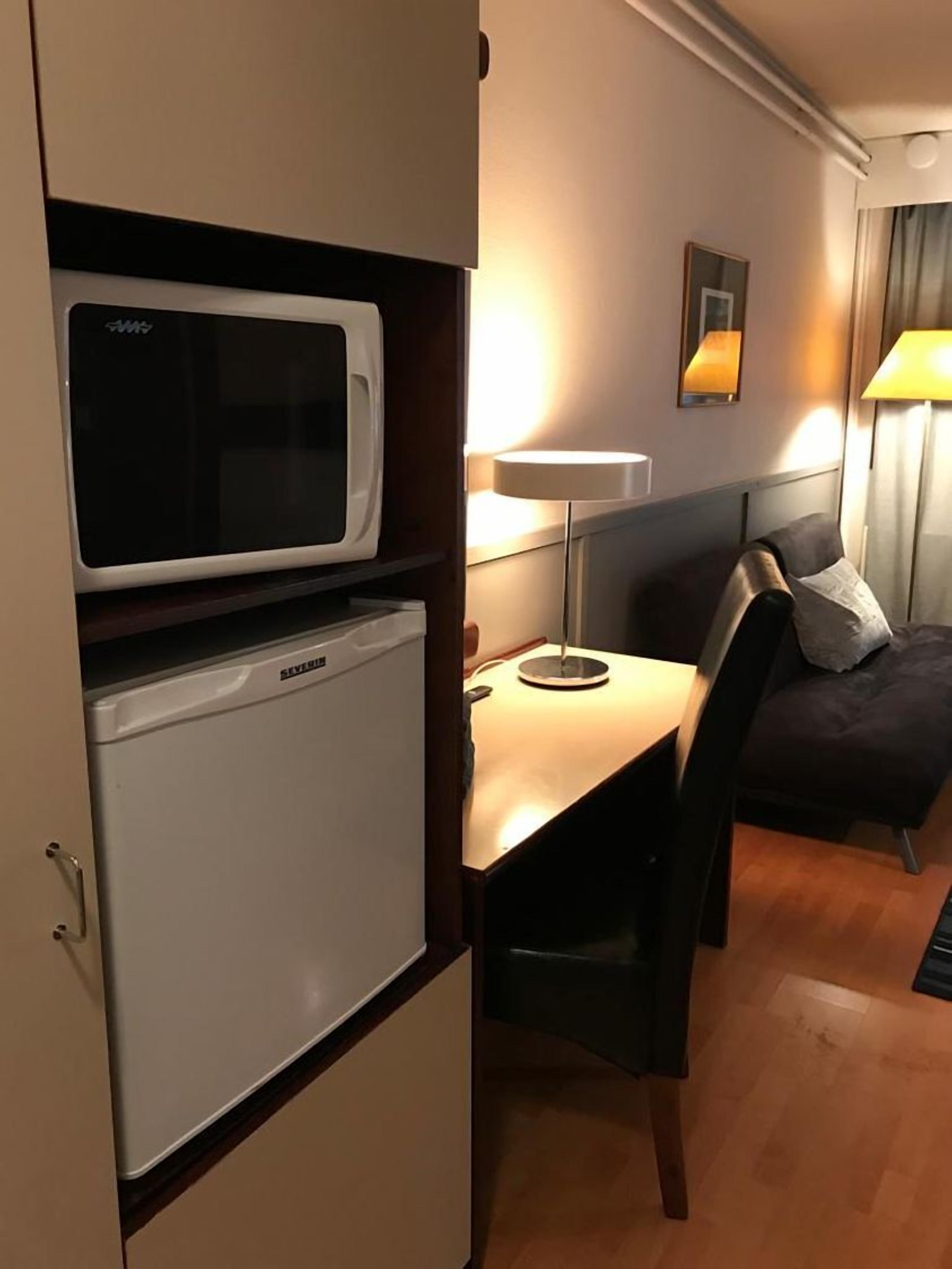 Uma kitchenette compacta com micro-ondas e frigorífico, uma pequena mesa de jantar com cadeiras, um abajur e um sofá de cor escura.