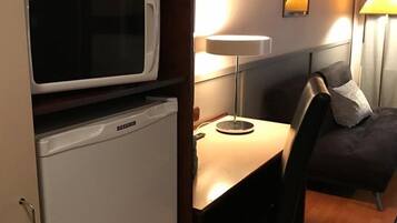 Uma kitchenette compacta com micro-ondas e frigorífico, uma pequena mesa de jantar com cadeiras, um abajur e um sofá de cor escura.
