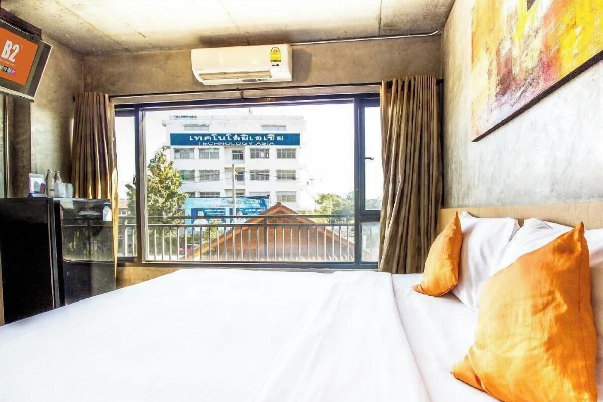 Quarto de hotel com cama, almofadas, janela com cortinas, ar condicionado e vista de um edifício.