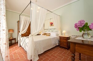 Quarto com cama de dossel, mesa de cabeceira com abajur, mesa de apoio de madeira e um vaso com flores.