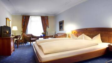 Quarto de hotel com uma cama grande, uma escrivaninha com televisão, duas cadeiras, um abajur e uma janela com cortinas.