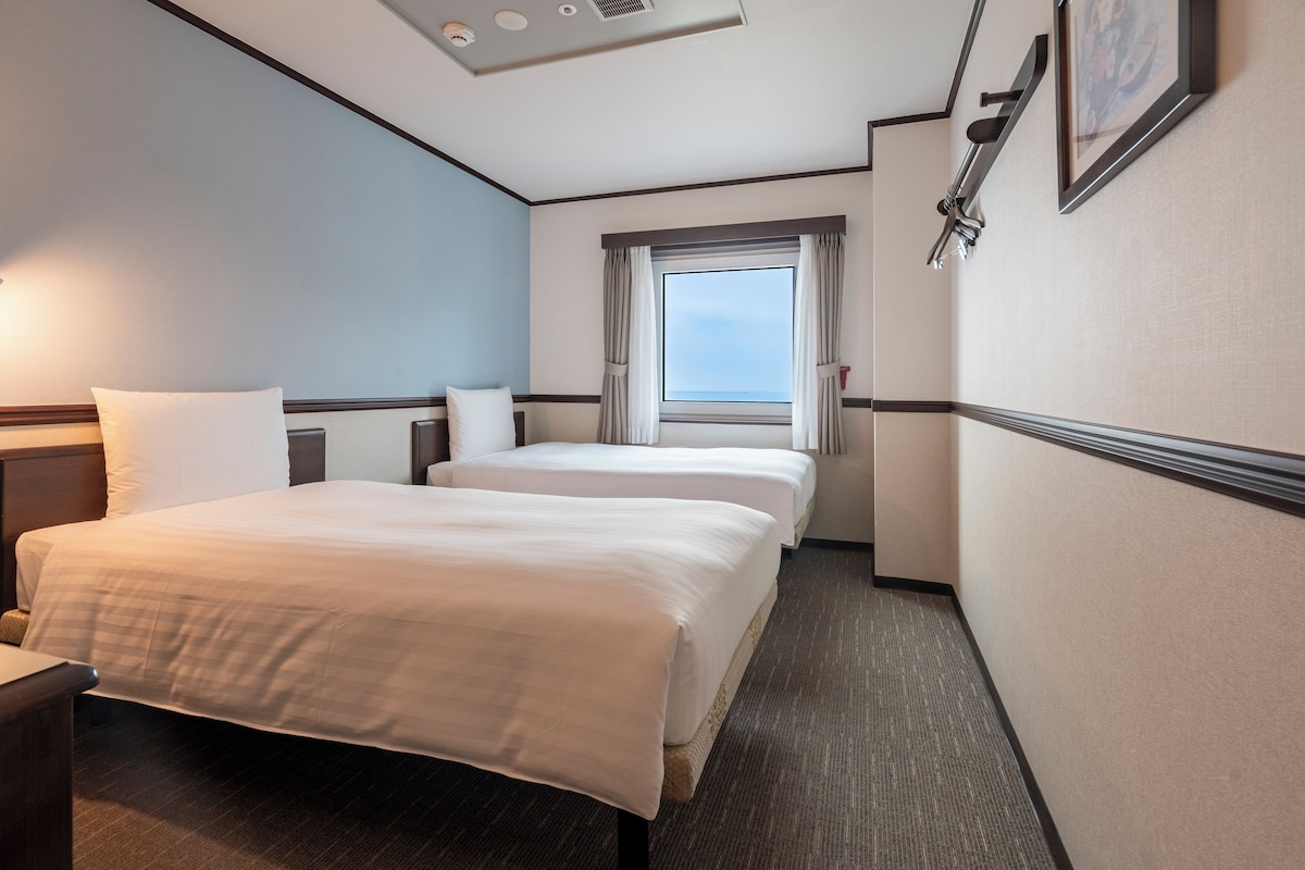 Quarto de hotel com duas camas, janela com cortinas, cabeceira e luminária fixada na parede.