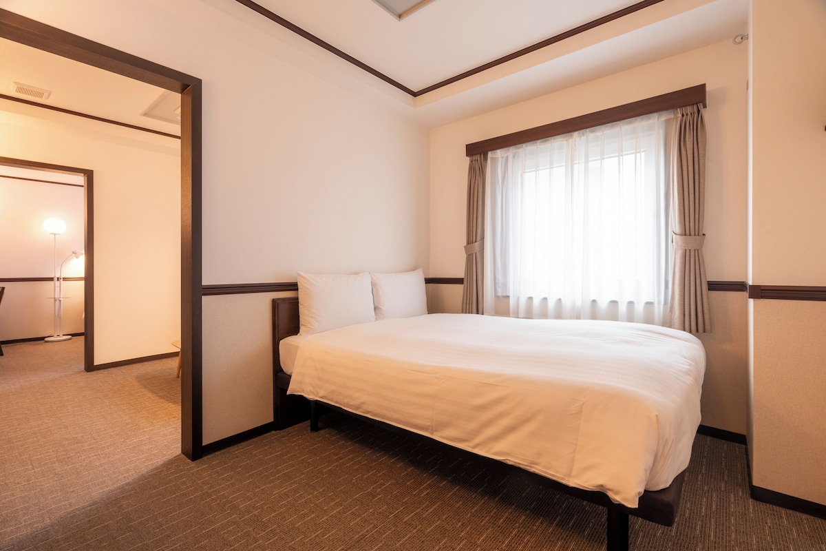 Quarto de hotel com uma cama grande, uma mesa de cabeceira, uma janela com cortinas e uma porta que dá para outro quarto.