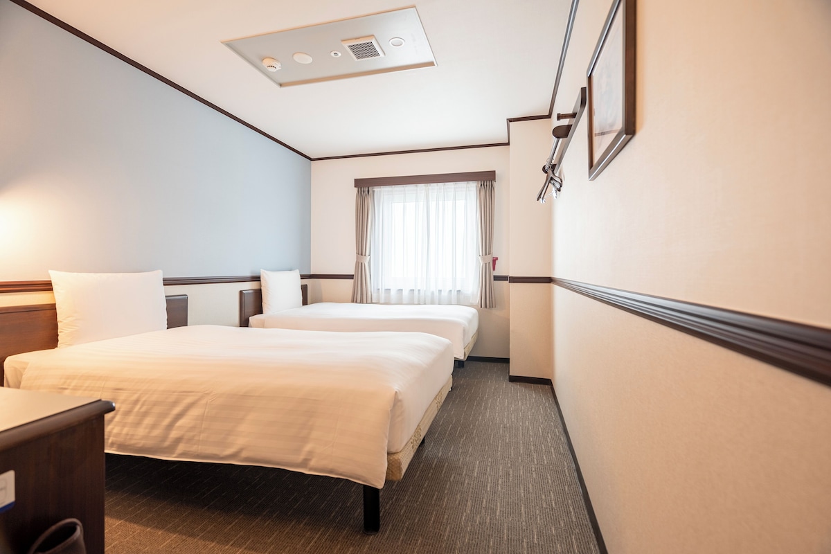 Quarto de hotel com duas camas, uma escrivaninha, uma cadeira, janela com cortinas e um quadro na parede.
