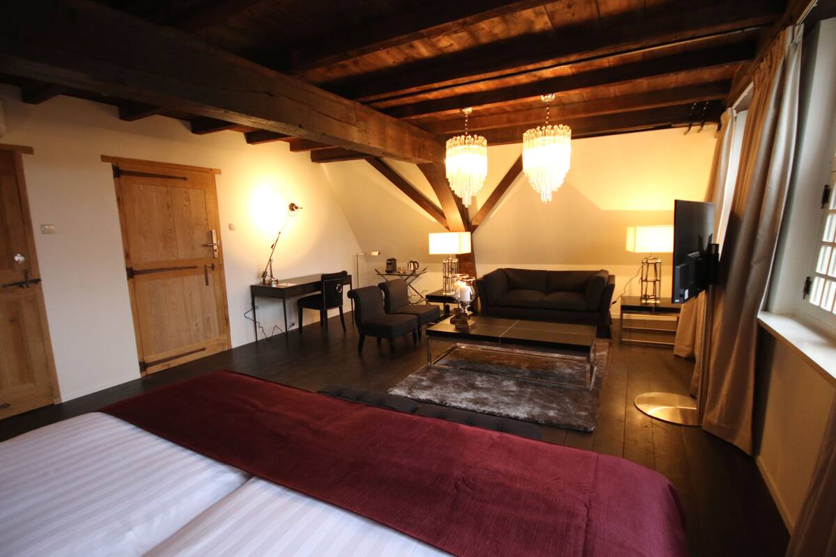 Quarto aconchegante com cama, sofá e mesa de jantar. Há lustres pendurados no teto e uma porta de madeira.