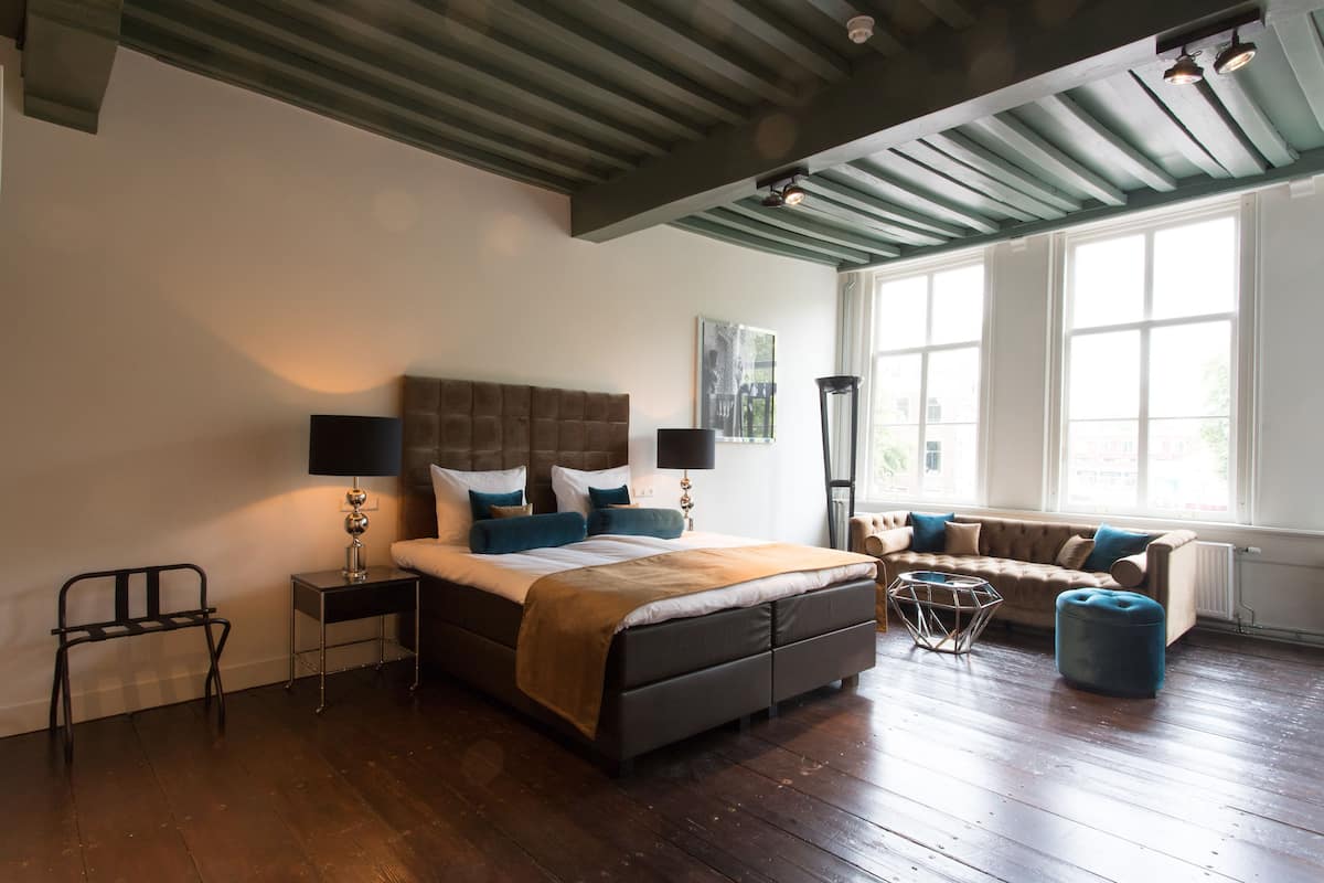 Quarto com cama grande, mesas de cabeceira, um banquinho e janela com luz natural.