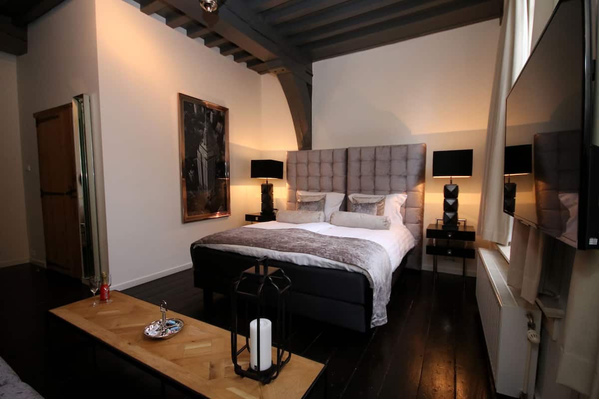 Quarto com cama grande, duas mesas de cabeceira com abajures, um criado-mudo de madeira, um quadro na parede e um radiador.